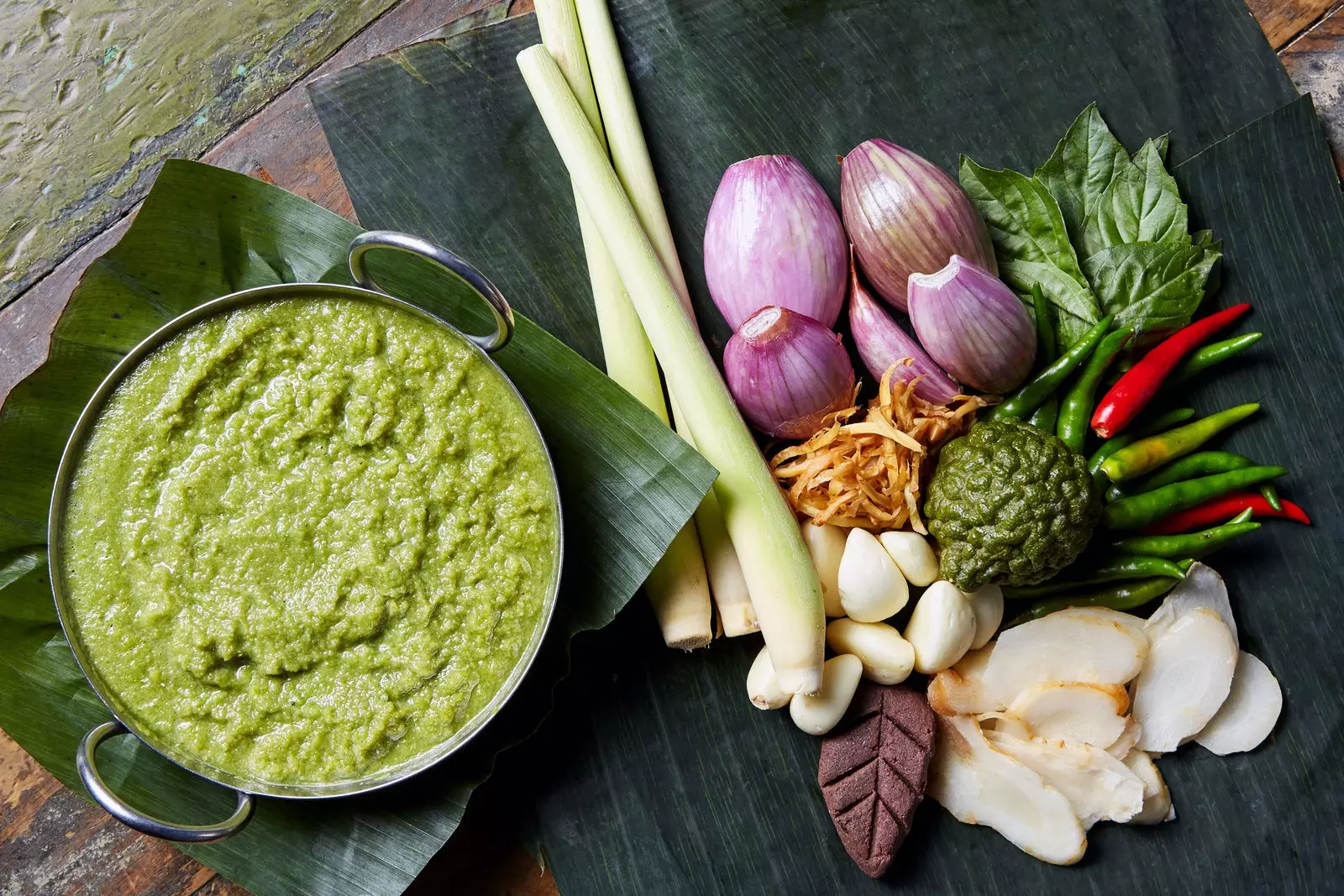 Foto von grünem Curry neben Zitronengras, Thai-Basilikum, Garnelenpaste, Kaffir-Limette, Ingwer, Knoblauch und Schalotten auf einem Bananenblatt.