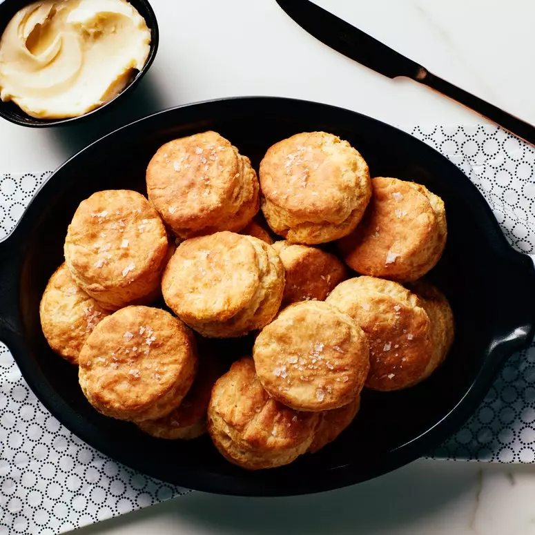 'n Hoop skilferige gelamineerde koekies op 'n swart skottel langs 'n klein skottel heuningbotter.