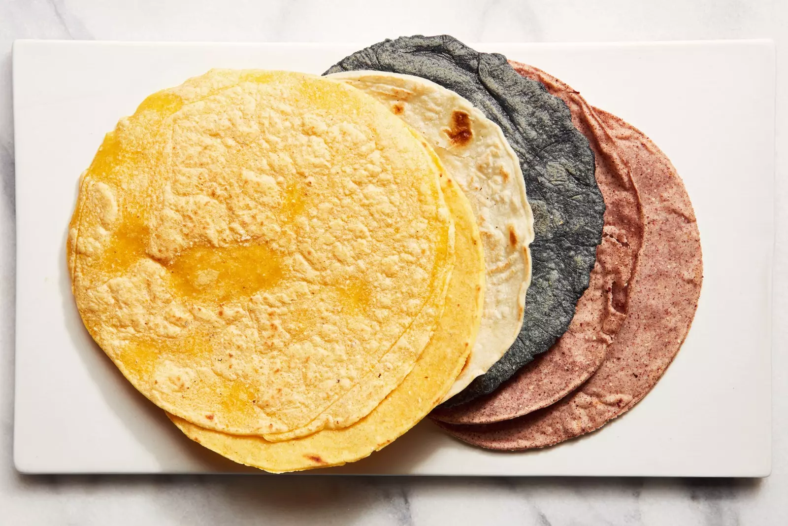 Fotografija žutih bijelih plavih i crvenih kukuruznih tortilja na mramornom tanjuru za posluživanje.