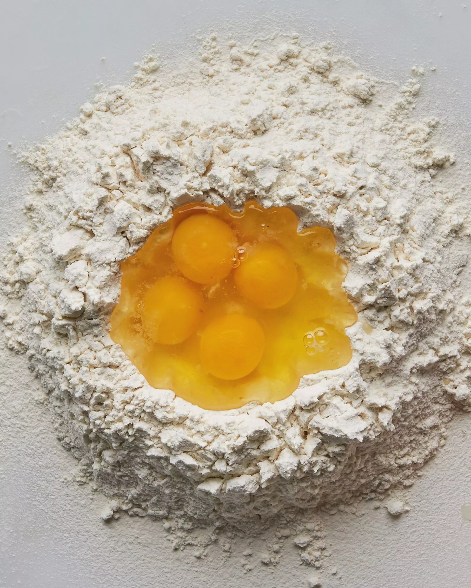 Zdjęcie czterech jajek rozbitych w studzience z mąką na marmurowym blacie.