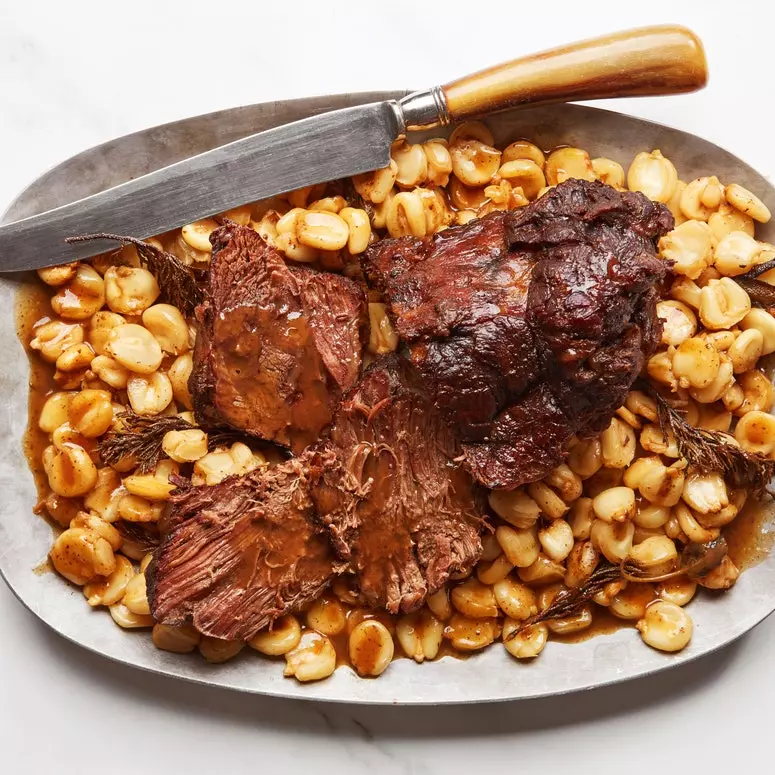 Foto Cedar Braised Bison di atas pinggan hidangan yang dikelilingi oleh hominy dan pisau ukiran.
