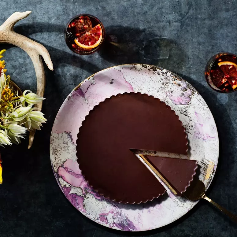 გიგანტური შოკოლადის არაქისის კარაქის ჭიქა ვარდისფერ თეფშზე ორი სასმლის ჭიქა და ყვავილები მუქი ნაცრისფერ ფონზე.