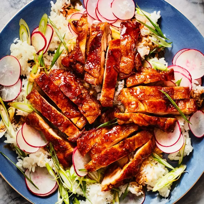 Teriyaki csirke kék tányéron fehér rizsszeletekre vágott retekkel és szeletelt mogyoróhagymával.