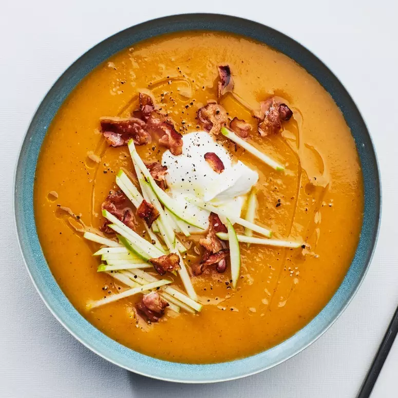 Unha cunca azul de sopa de cabaciña e mazá cuberta con touciño de crema de leite e mazás en rodajas finas.