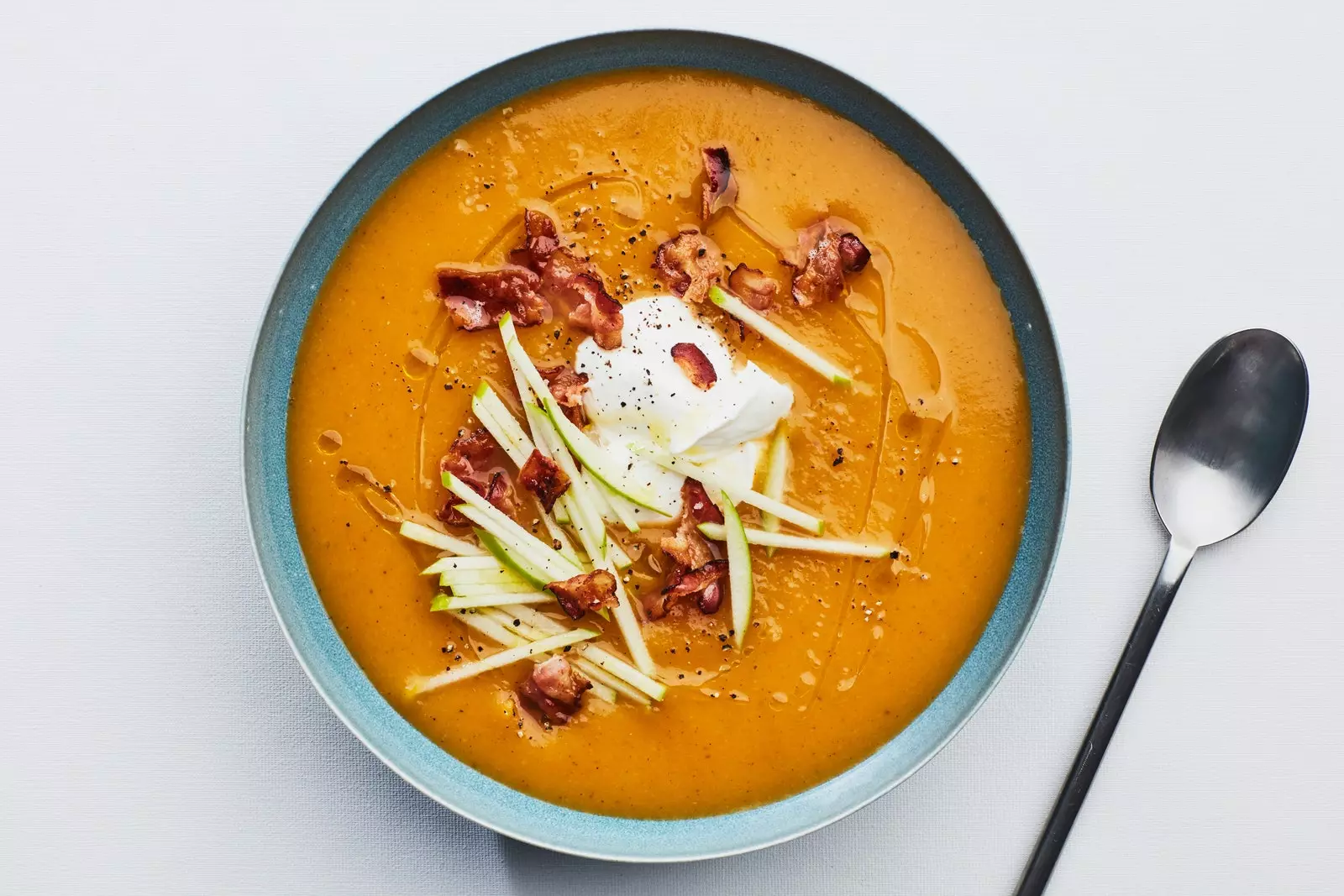 Un bol bleu de soupe à la courge musquée et aux pommes garni de bacon à la crème sure et de pommes tranchées finement.