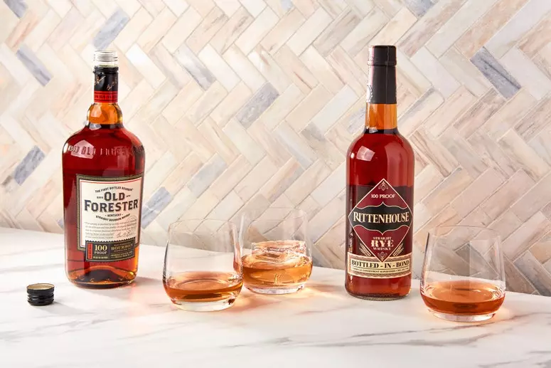Fotografie cu o sticlă de Old Forester Straight Bourbon și o sticlă de Rittenhouse Straight Rye, cu trei pahare, pentru turul nostru cu cel mai bun whisky.