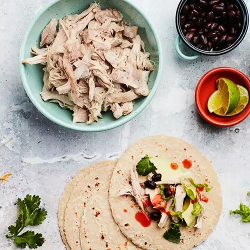 Eine Schüssel mit zerkleinertem Hähnchen neben einem Haufen Tortillas, eine Schüssel mit gehackten Tomaten, eine Schüssel mit schwarzen Bohnen und ein kleines Gericht...