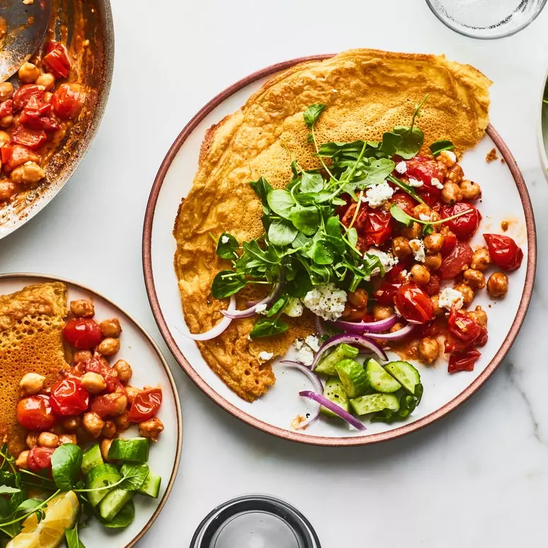 Olivenueleg-baséiert Chickpea flatbread tëscht zwee Placke mat geplatzt Tomatenzooss a Puppelchen gréng verdeelt.