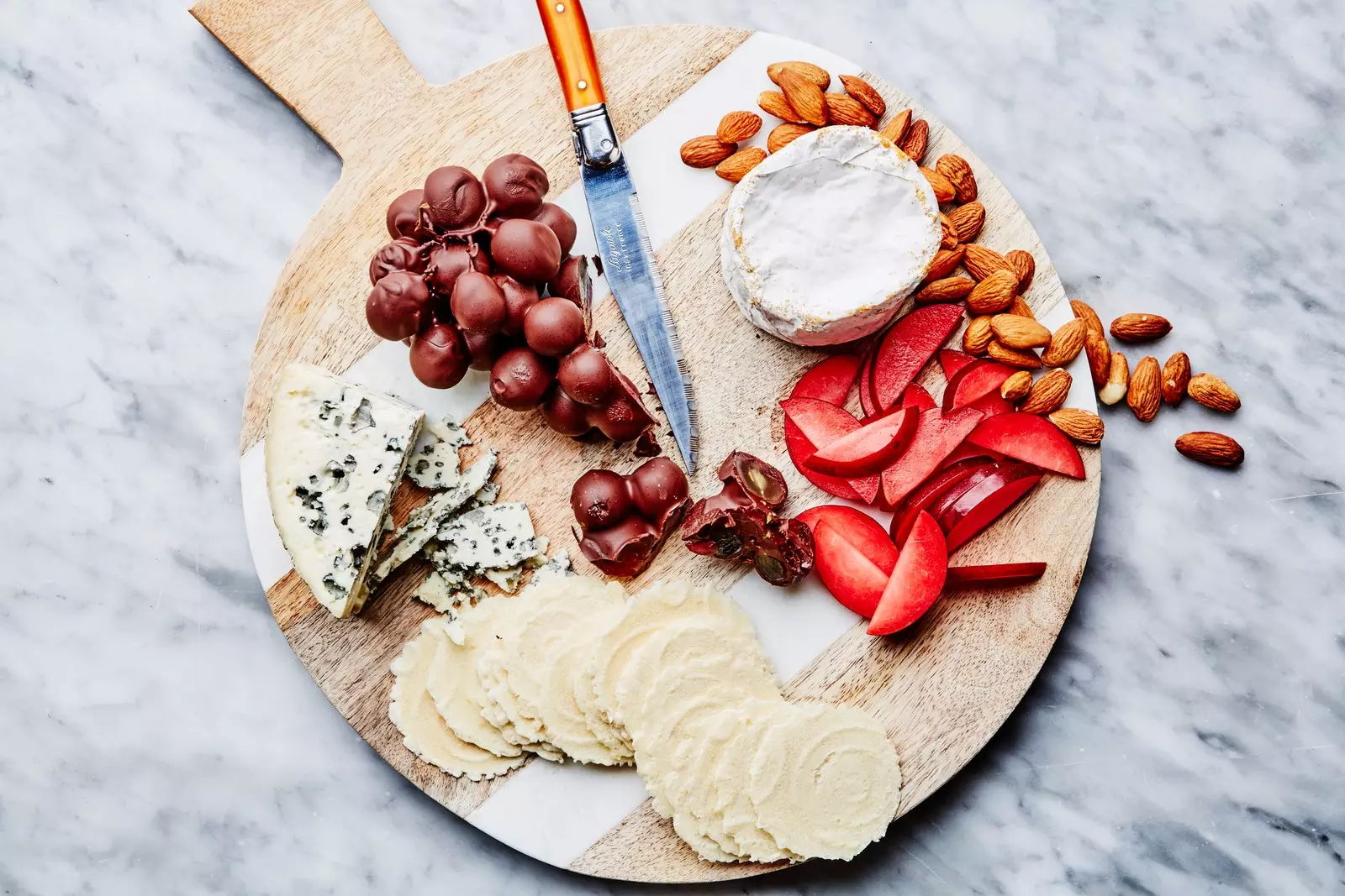 Uma placa de queijo redonda com crackers de uvas fatias de ameixas e alguns queijos com uma faca.