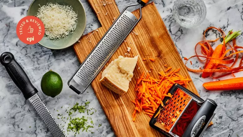 Litrato sa tulo ka lain-laing graters sa usa ka marmol countertop uban sa Parmesan keso, carrots, ug apog.