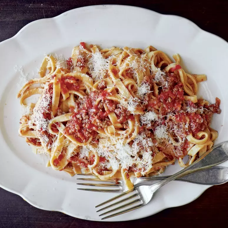 Bolonhesa com tagliatelle coberto com parmesão em uma travessa branca com garfos de prata.