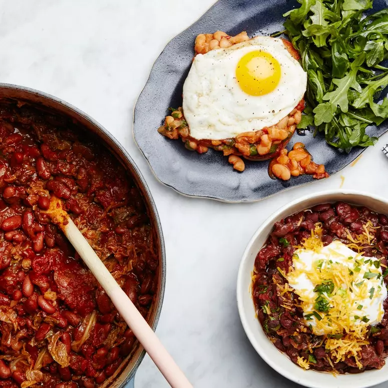 Bagte bønner i en hollandsk ovn med en træske. Ved siden af er en tallerken og skål både fyldt med baked beans og toppet...