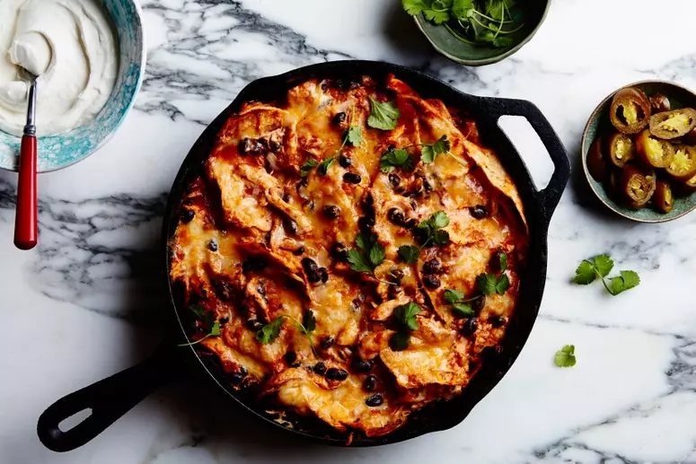 Una paella negra de ferro colat amb enchiladas de pollastre i bols de tomàquets cherry a la meitat, fulles de coriandre i crema agra al costat.