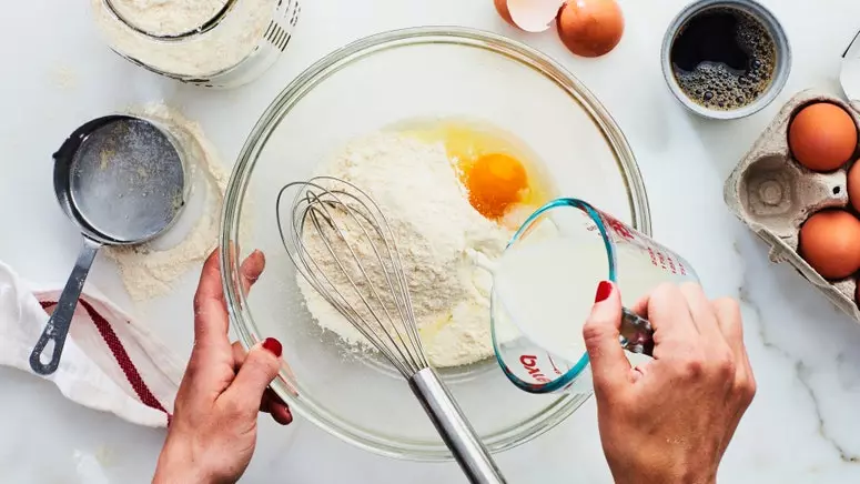 En översiktsbild av någon som häller mjölk i en skål med en visp, ett ägg och torra ingredienser till pannkakor.