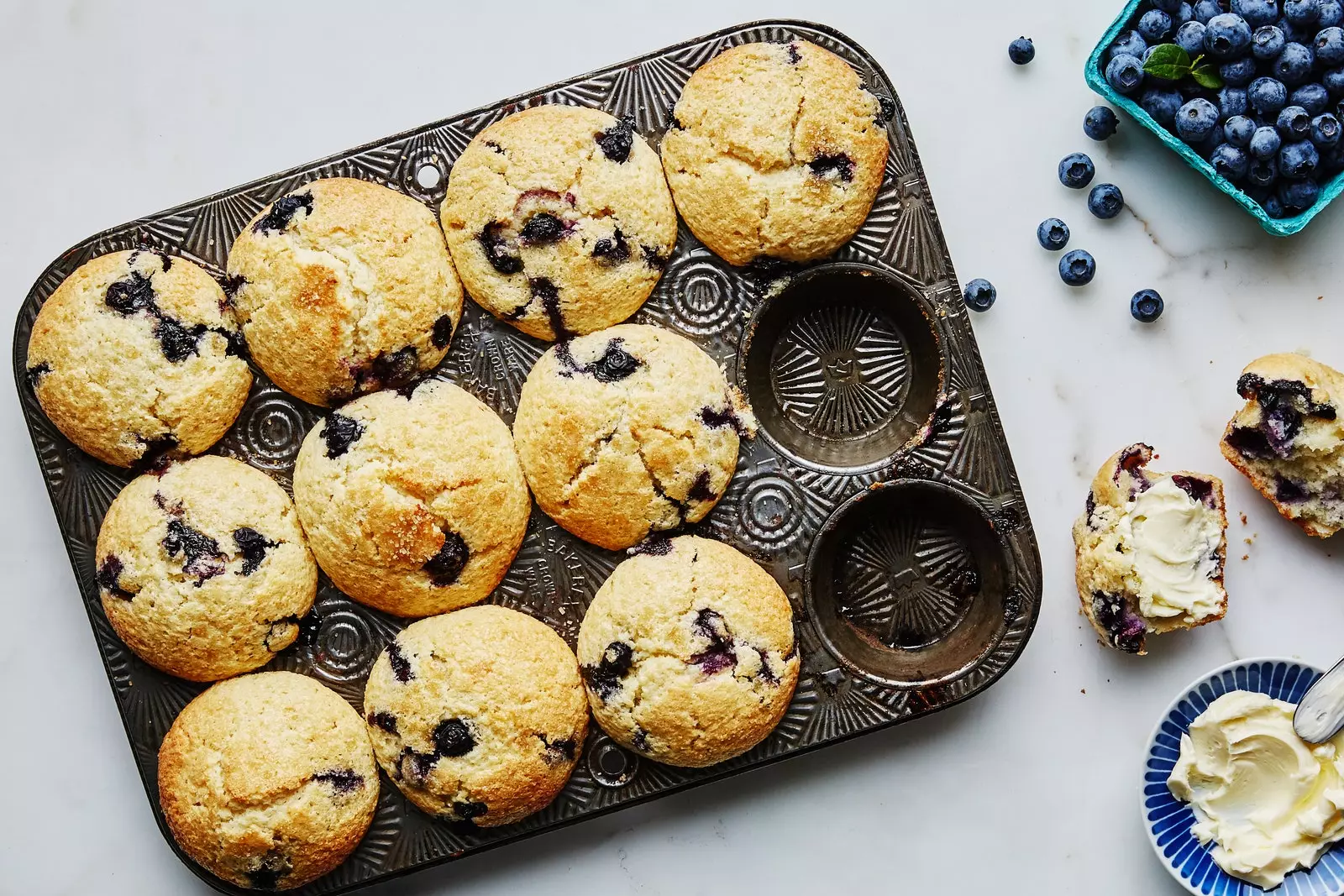 Kurasa hejuru yubururu bwa blueberry mumabati ya kera hamwe na muffins ebyiri zavanyweho iruhande rwa pint yubururu bushya hamwe na ...