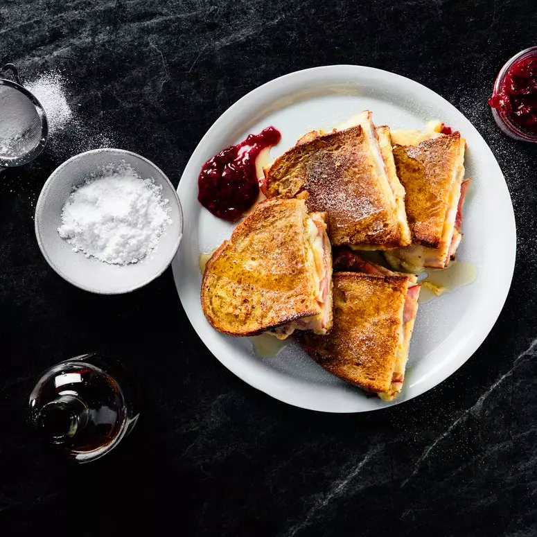 Roti lapis Monte Cristo di atas piring putih di atas meja hitam.