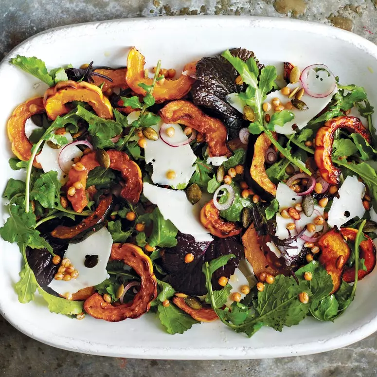 Arugula na nilagyan ng roasted acorn squash pumpkin seeds at keso sa puting rectangular serving dish.