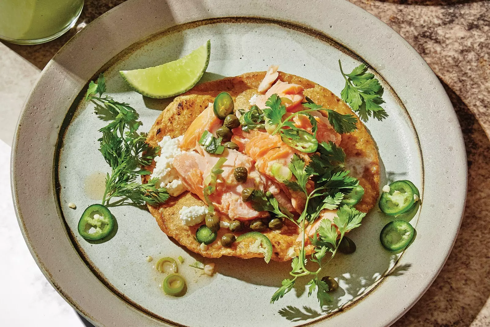 Sült lapos tortilla kerámia tányéron, tetején pelyhes rózsaszín hallal, szeletelt jalapeños korianderrel és kapribogyóval.