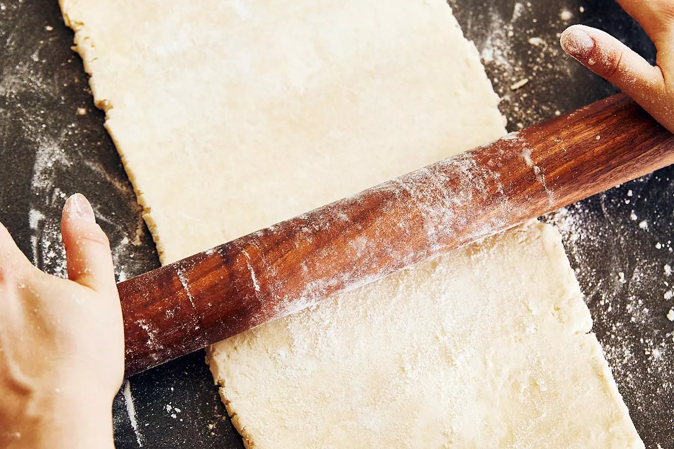 Un rodillo de amasar de madera de cerezo y las manos de una mujer joven extendiendo un trozo de pastel doblado.