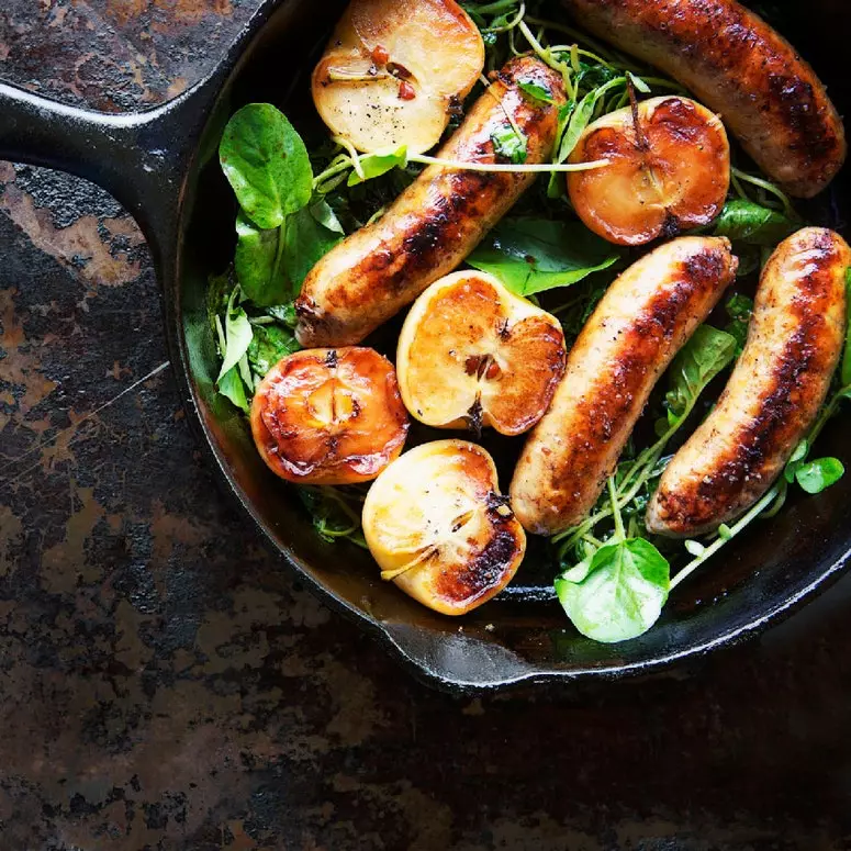 Saucisses poêlées dans une poêle en fonte avec des pommes coupées en deux et du cresson frais.