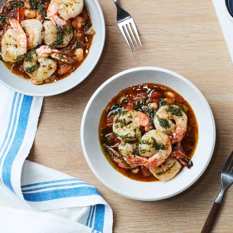 Foto udang dalam mangkuk dengan kaldu kacang putih dan tomat ditampilkan dengan garpu dan beberapa potong roti panggang.