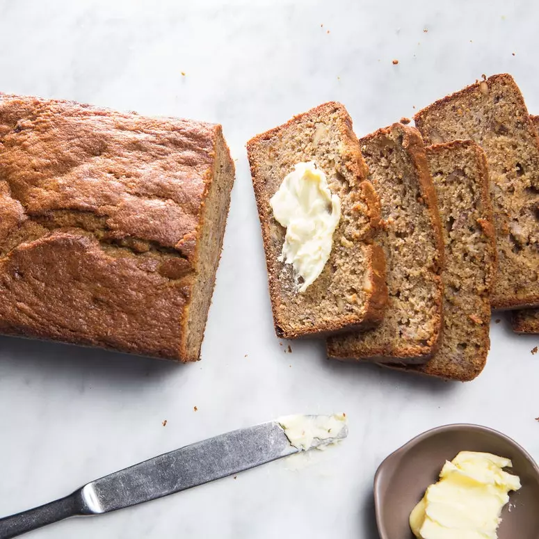 Et brød bananbrød i halve skiver på en marmorflate med en tallerken med mykt smør ved siden av.