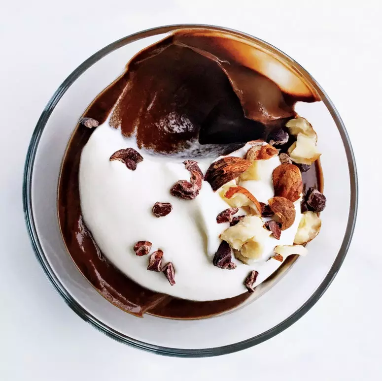 Púding d'alvocat de xocolata en un bol de vidre cobert amb un rajolí de grans de cacau de nata i una mica de fruits secs.