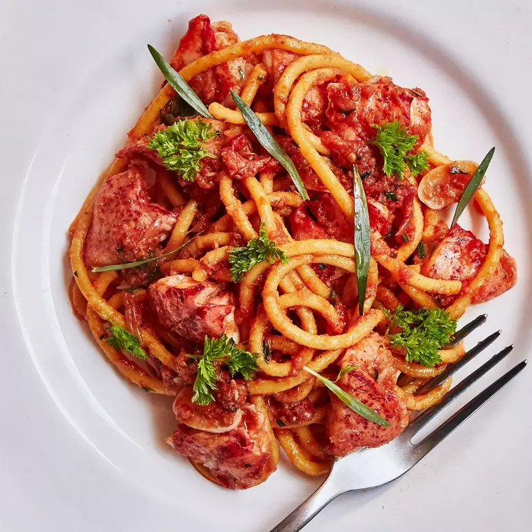 叉子与意大利面交织在一起，意大利面在红色酱汁中加入龙虾块和香草，放在白色的褶边盘子上。