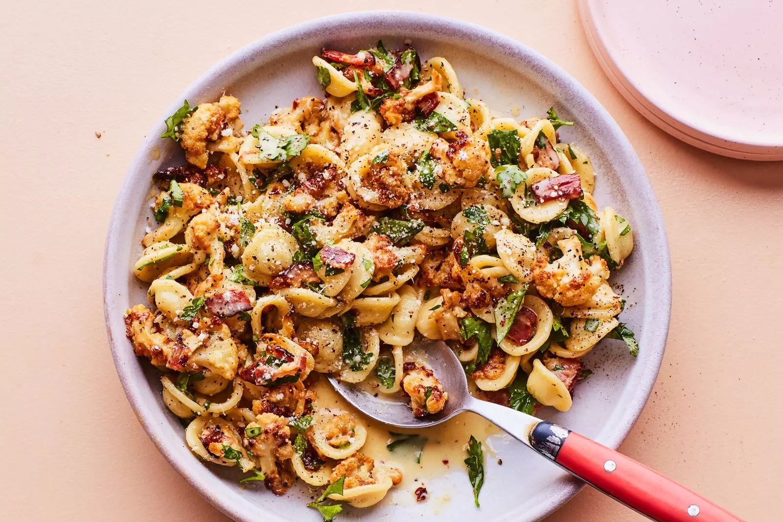 Blomkool carbonara gemaak met orecchiette op 'n pienk bord met 'n rooihandvatsellepel.