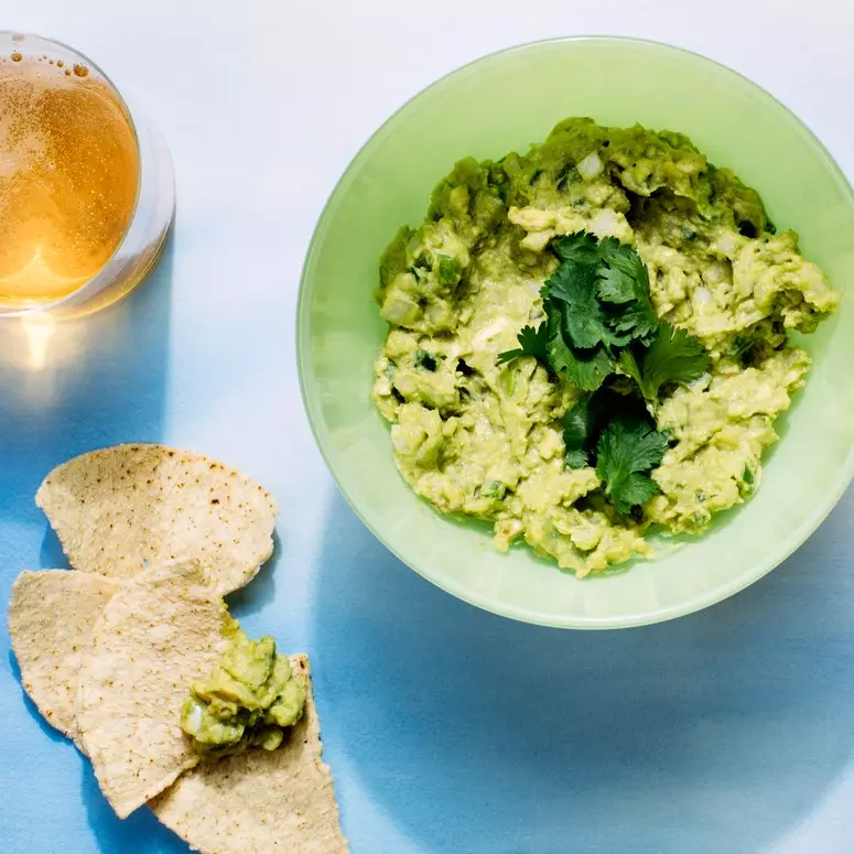 L-aqwa riċetta tal-guacamole fi skutella ħadra mimlija cilantro frisk. Fuq il-ġenb hemm skutella tortilla chips u...