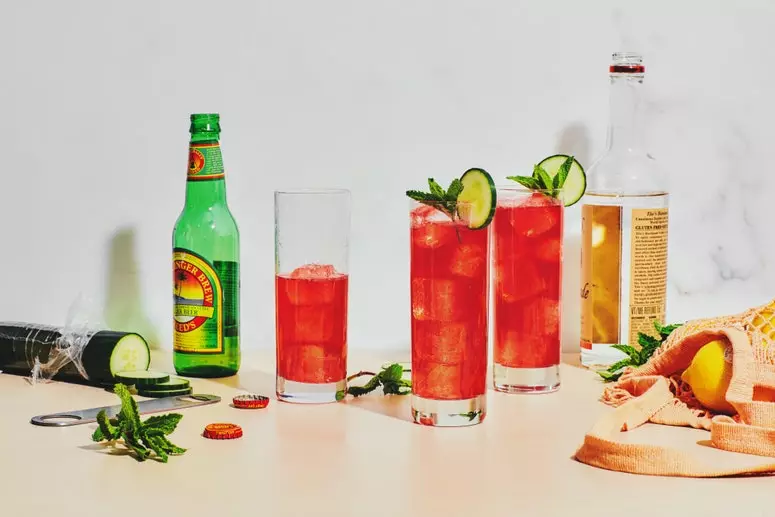 Drie levendige rode cocktails in hoge, met ijs gevulde glazen gegarneerd met komkommer en munt, naast flessen gemberbier en wodka, plus een gesneden komkommer en een boodschappentas met netten.