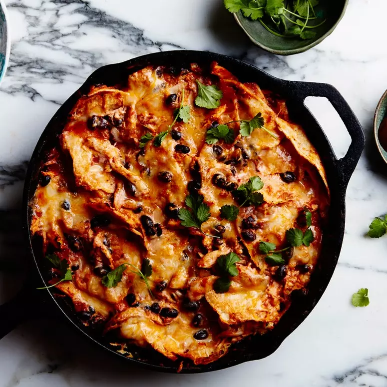 Una padella di ghisa nera con enchiladas di pollo e ciotole di pomodorini tagliati a metà foglie di coriandolo e panna acida accanto...