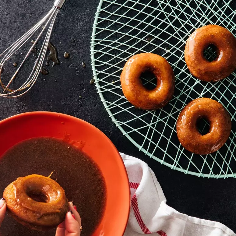 Vier appelcider-donuts op een koelrek met blauwe cirkel met een kopje koffie en garde ernaast. In de linkerhoek...