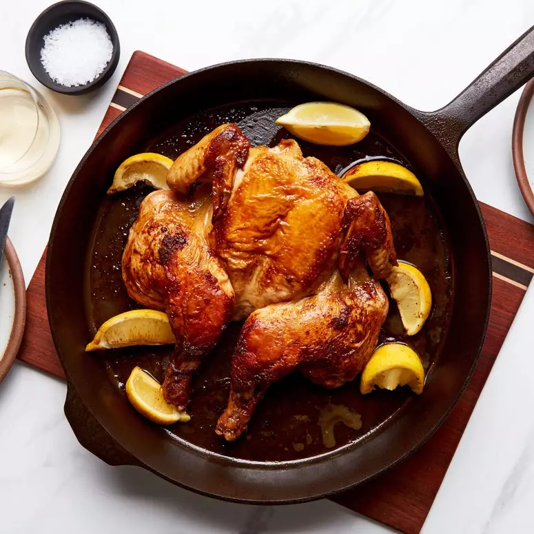 Foto van een hele kip in een gietijzeren koekenpan omringd door schijfjes citroen op een houten snijplank met borden en...