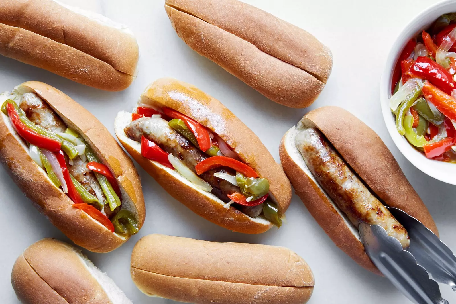 Foto de salsichas sendo colocadas em pães de cachorro-quente divididos com pimentão e cebola.