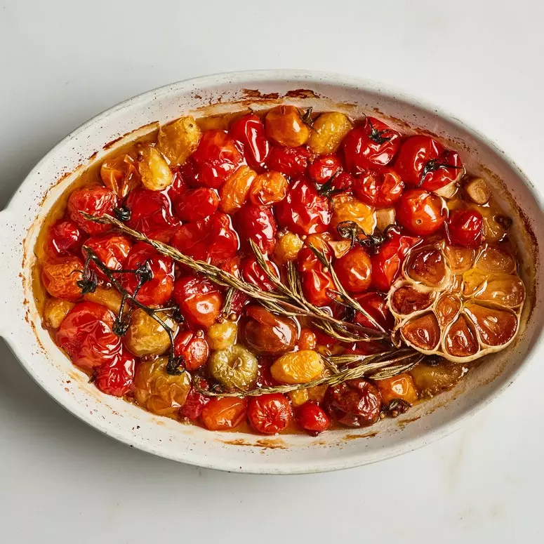 Pečene cherry rajčice s grančicom ružmarina i pola glavice češnjaka u bijeloj ovalnoj posudi za pečenje na mramornoj površini.