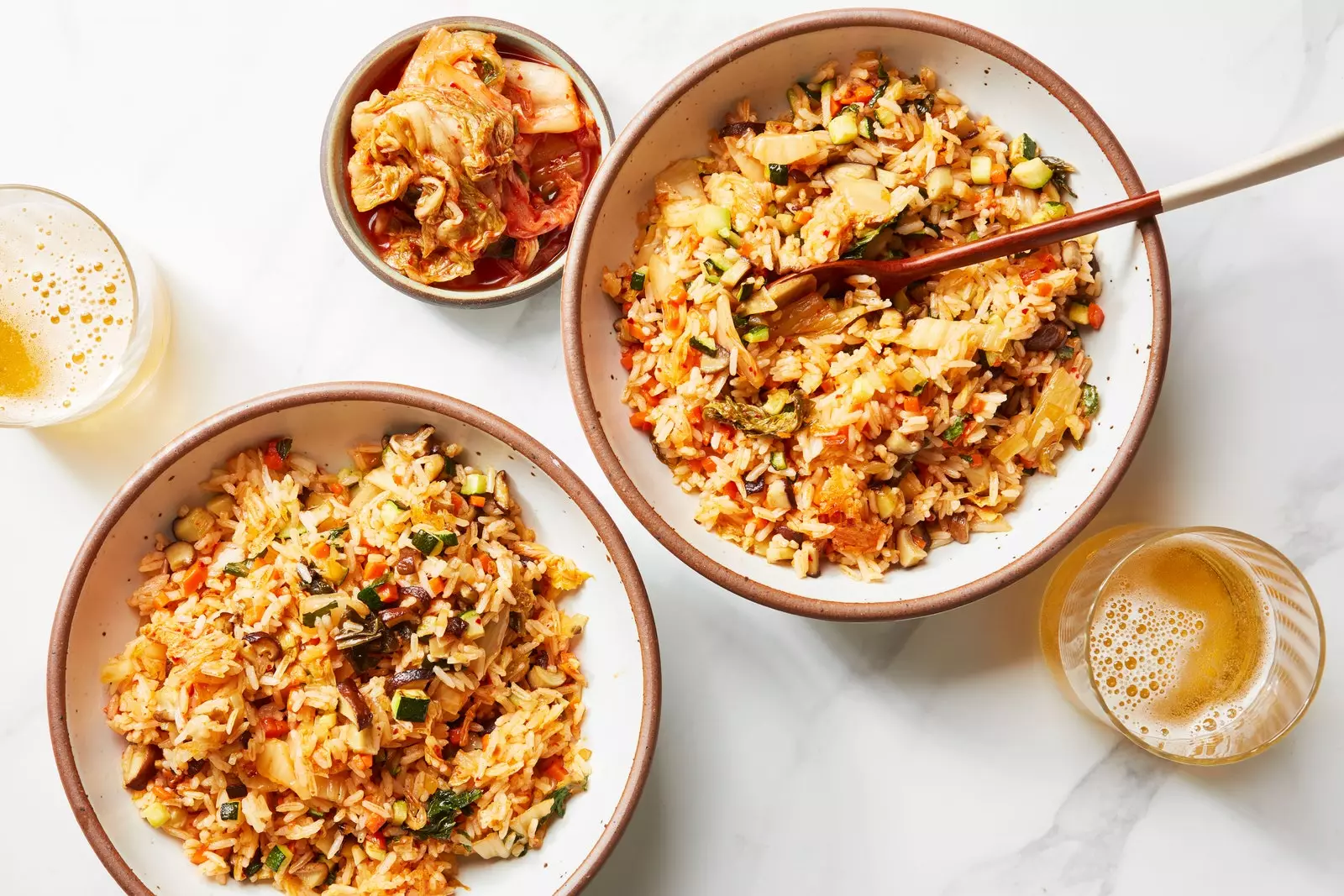 Rong mangkok Nasi Goreng Kimchi karo mangkok kimchi lan rong gelas bir ing sisih pinggir.