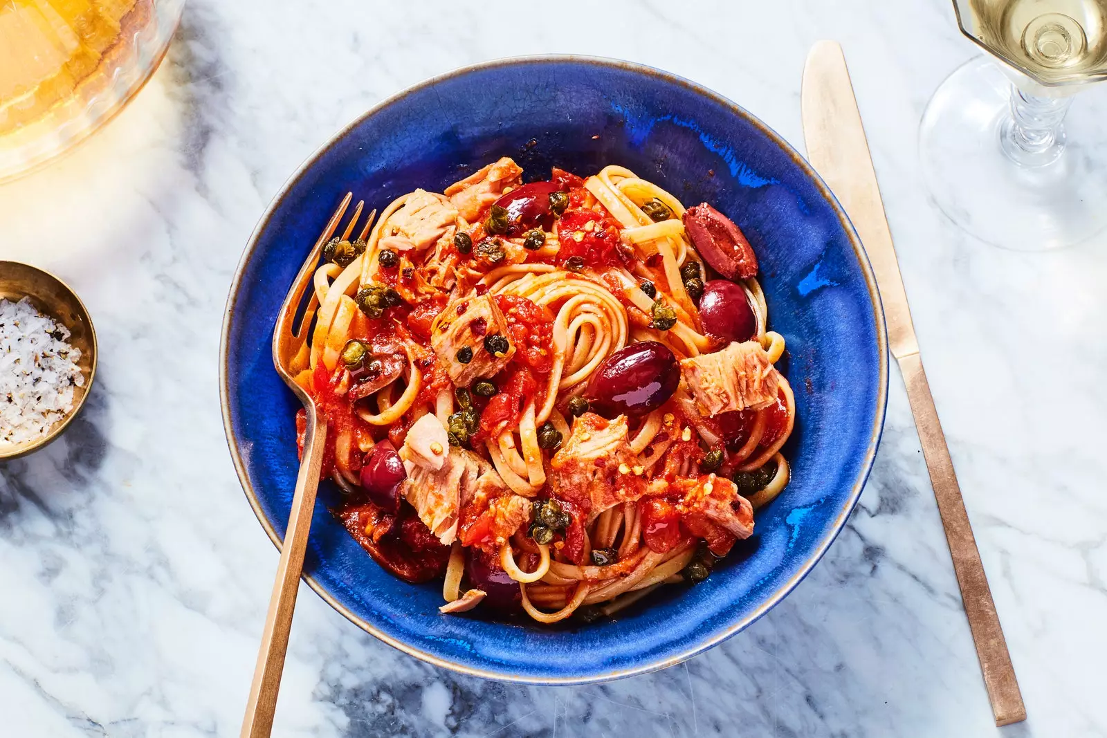 Pasta Puttanesca an enger bloer Schuel mat enger gëllener Gabel a Messer.