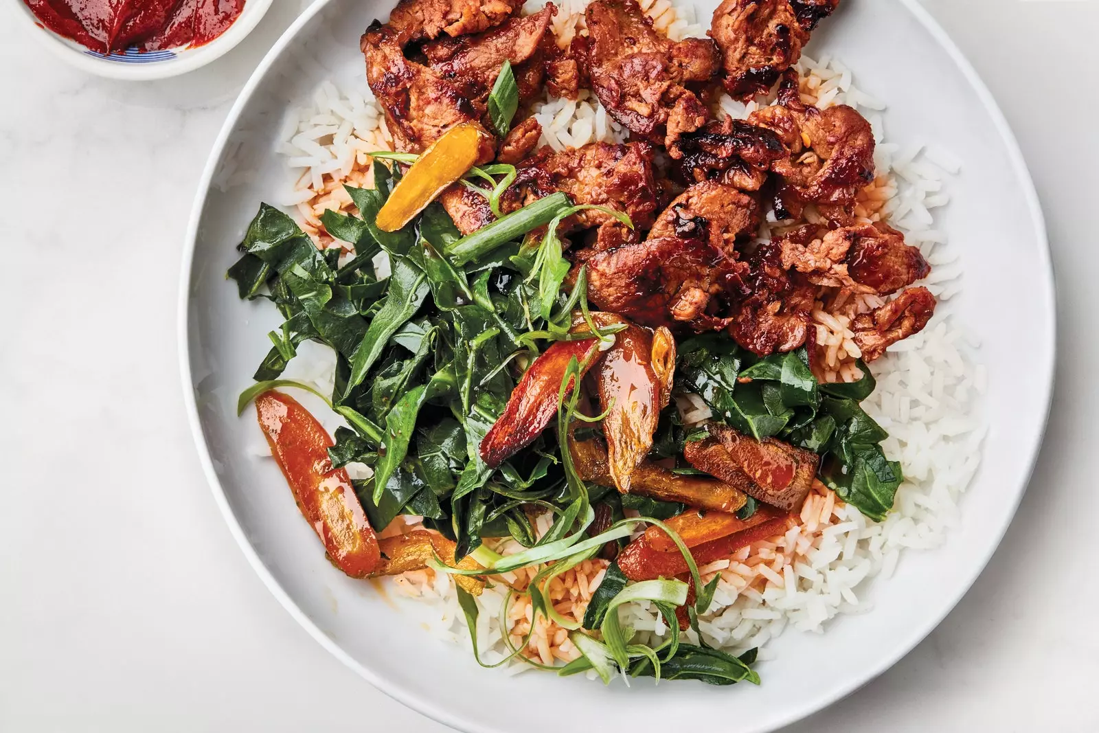 Isang malawak na mababaw na mangkok na puno ng steamed white rice na nilagyan ng glazed strips ng pork tenderloin at carrots na may tambak na...