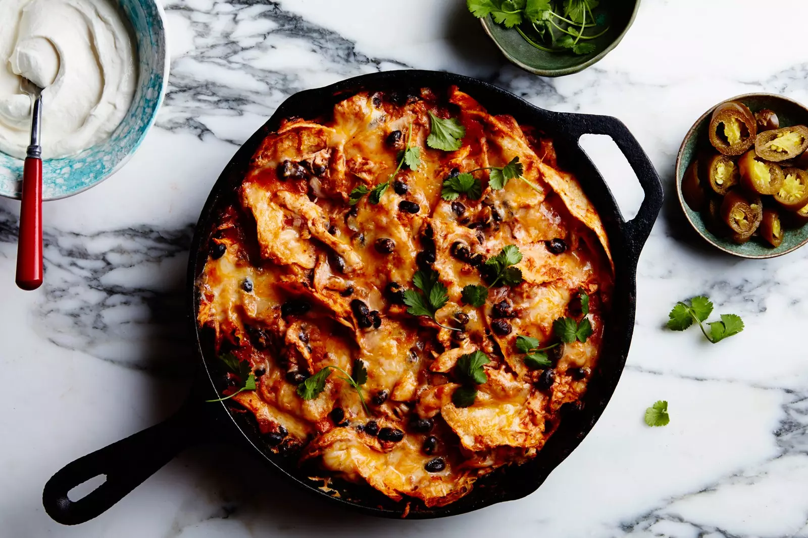 Isang black castiron skillet na may chicken enchilada at mga mangkok ng halved cherry tomatoes na dahon ng cilantro at sour cream sa susunod...