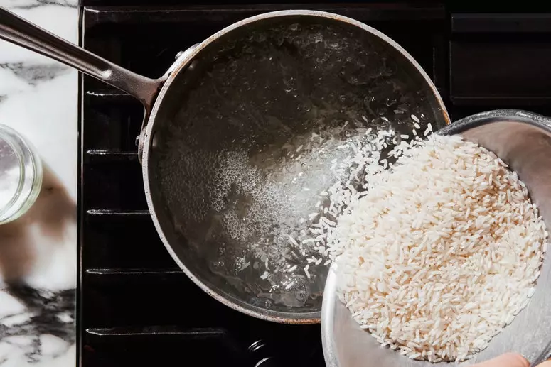 Panela de fogão com arroz sendo despejado em água fervente com sal ao lado.