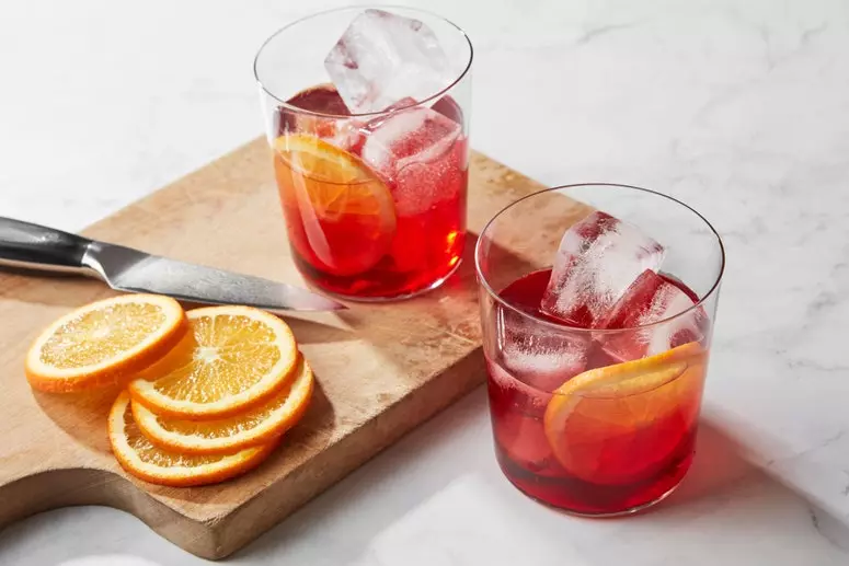 Фотография двух стаканов безалкогольного негрониса рядом с разделочной доской с дольками апельсина.