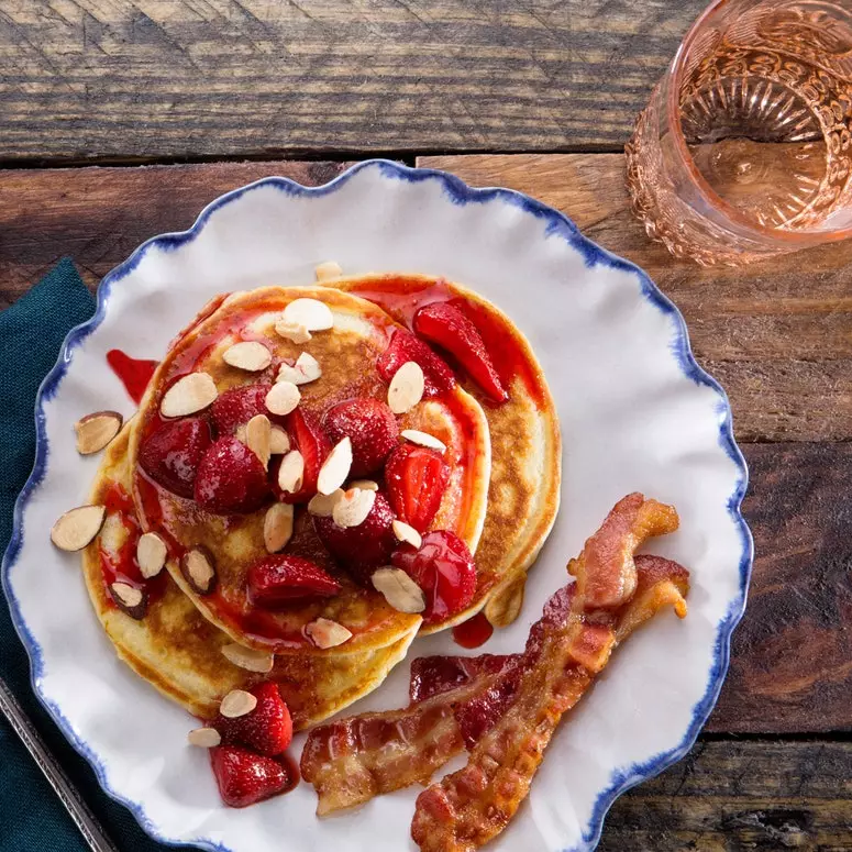 En tallerken pannekaker stablet høyt og toppet med saucy halverte jordbær og mandler med en side av bacon pluss en stor...