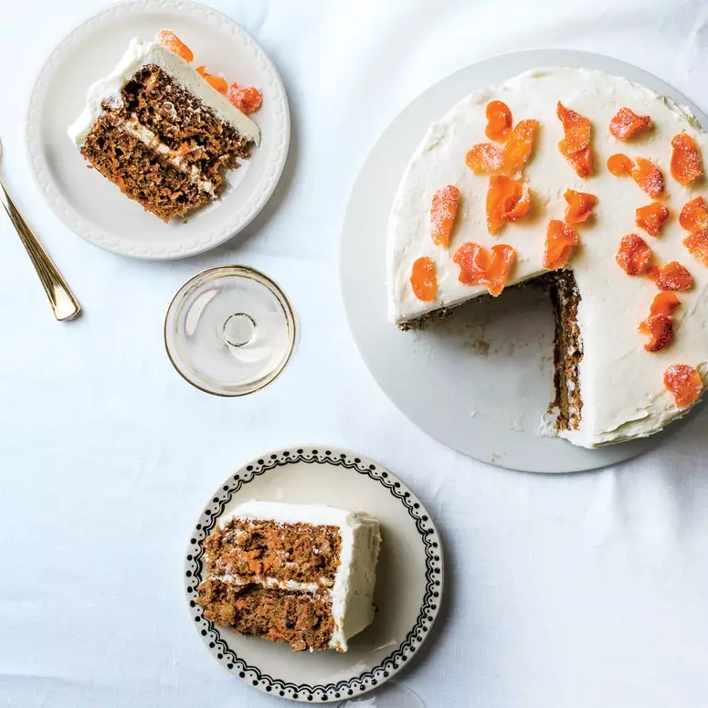 Layered carrot cake nga adunay cream cheese nga frosting nga gibutang sa ibabaw sa candied carrot coins sa usa ka serving dish nga adunay duha ka hiwa nga giputol...