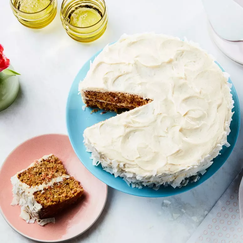 Císte cnó cócó cairéad dhá shraith le icing bán cnó cócó ar phláta gorm le slisne gearrtha uaidh.