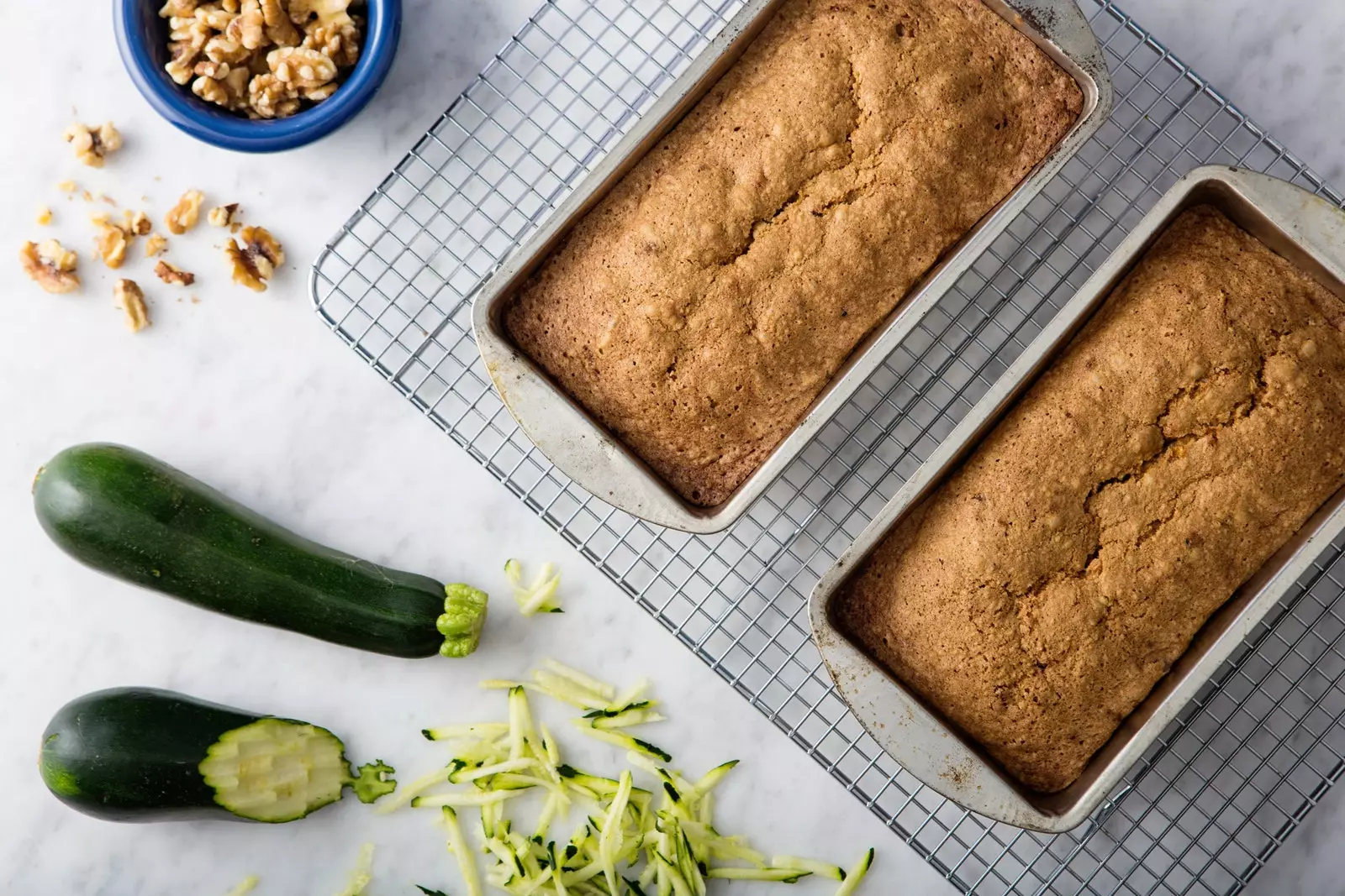 Due pagnotte di pane alle zucchine in teglie adagiate su una gratella con altre zucchine parzialmente sminuzzate e noci fuori...