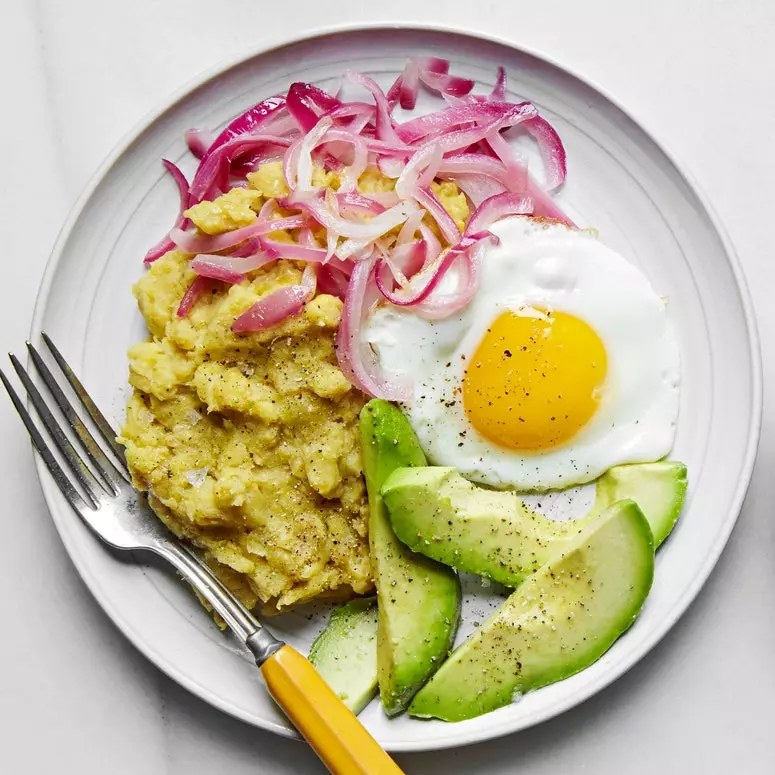 Foto af mosede plantains med skiver avocado, spejlæg og rødløg.