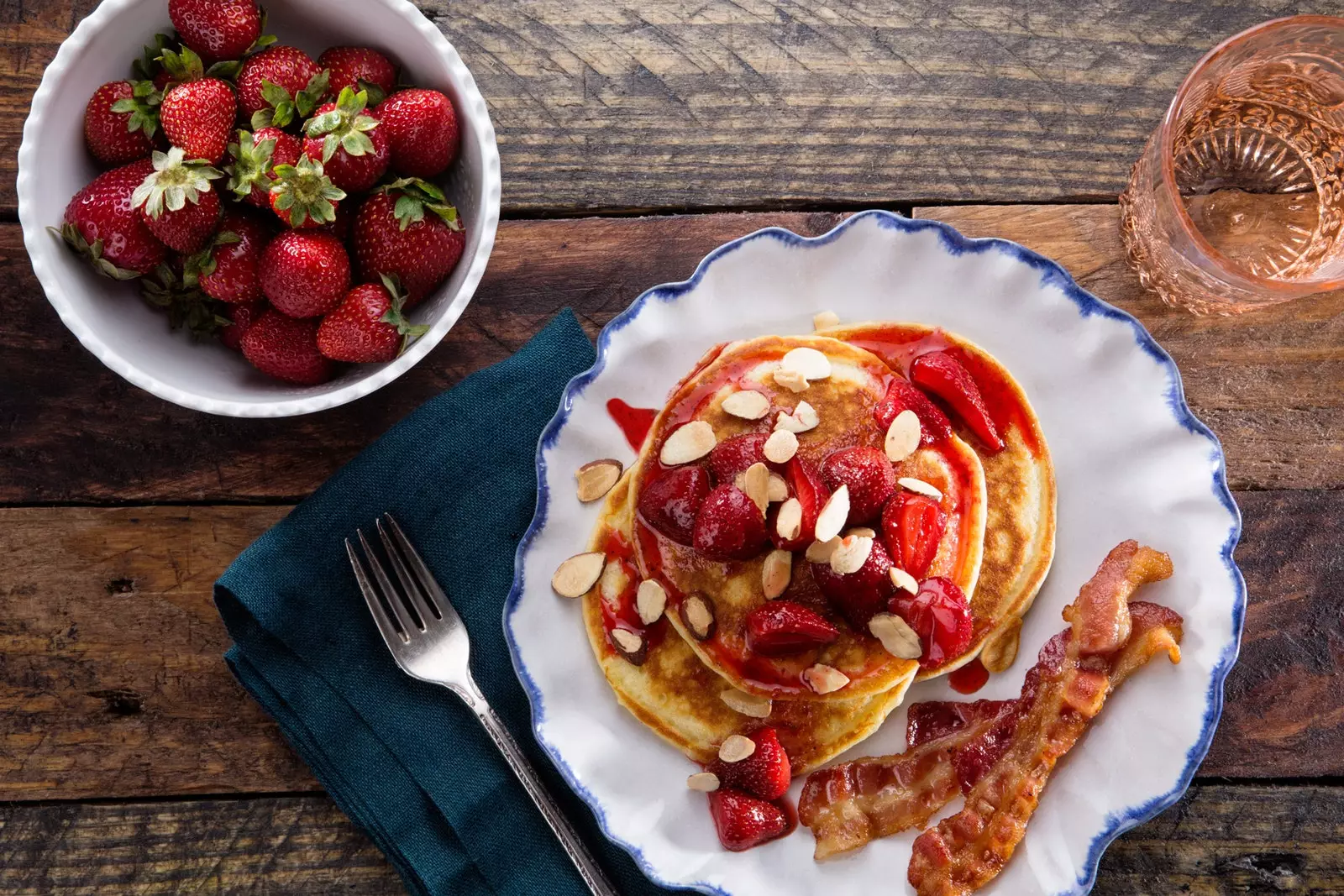 Un plato de panqueques apilados alto y cubierto con salsas de fresas cortadas por la mitad y almendras con una guarnición de tocino más un gran...