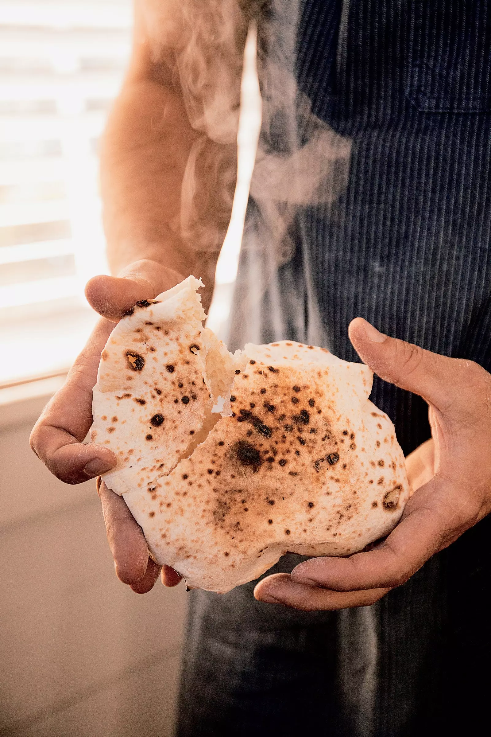 Dłonie Alona Shayi rozrywają gorącą pitę unoszącą się z chleba.
