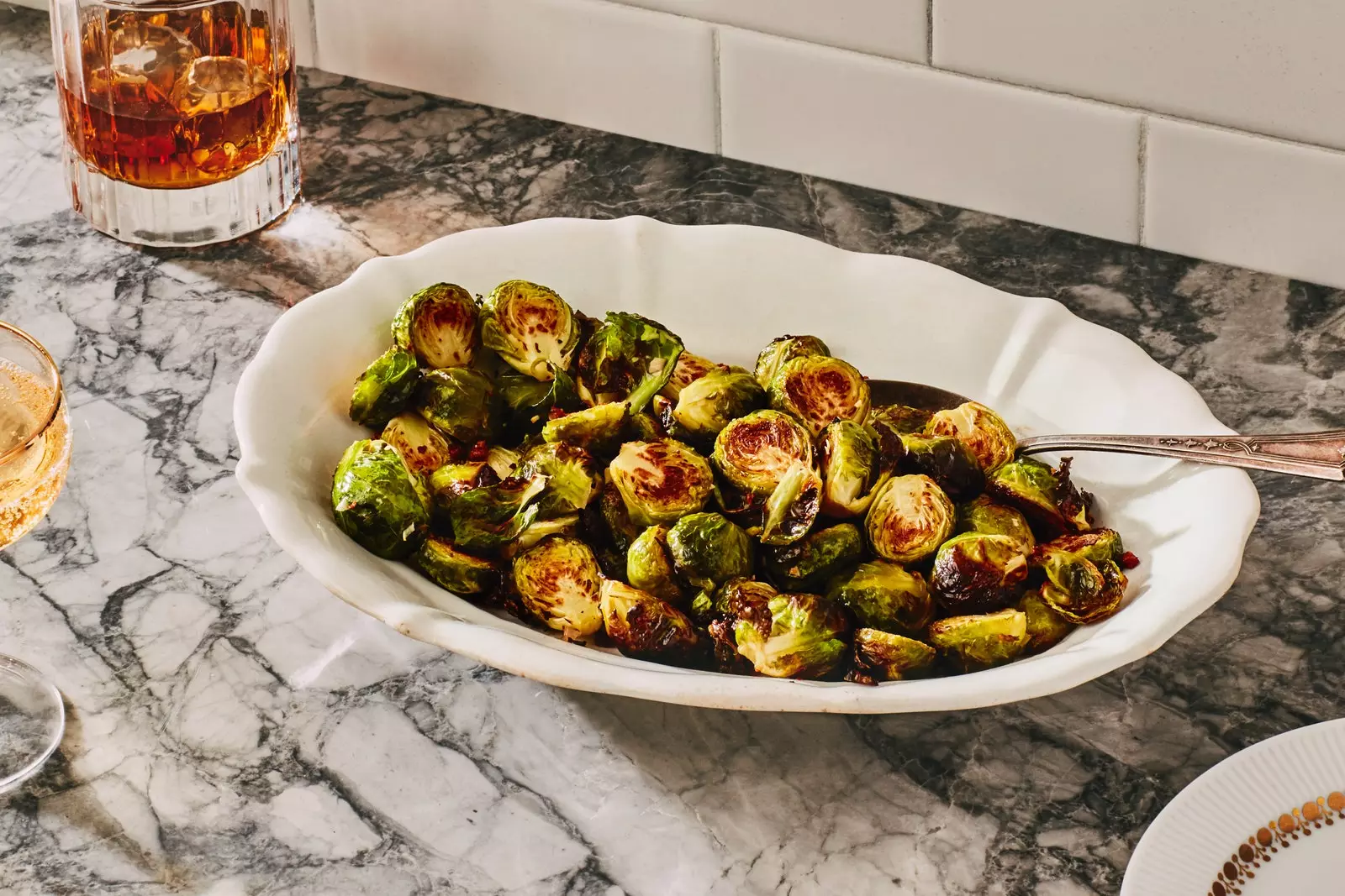 Ti yan Brussels Sprouts pẹlu ata ilẹ ati Pancetta ninu satelaiti pẹlu ṣibi ti o n ṣiṣẹ ati awọn ohun mimu ni ẹgbẹ